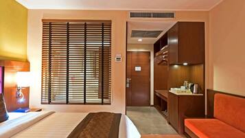 Quarto de hotel com uma cama, um sofá, um guarda-roupa de madeira, uma escrivaninha com chaleira e uma janela com persianas.