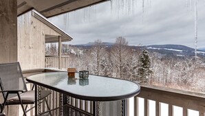 Uma varanda com mesa e cadeiras de vidro, com vista para uma paisagem nevada com árvores e montanhas.