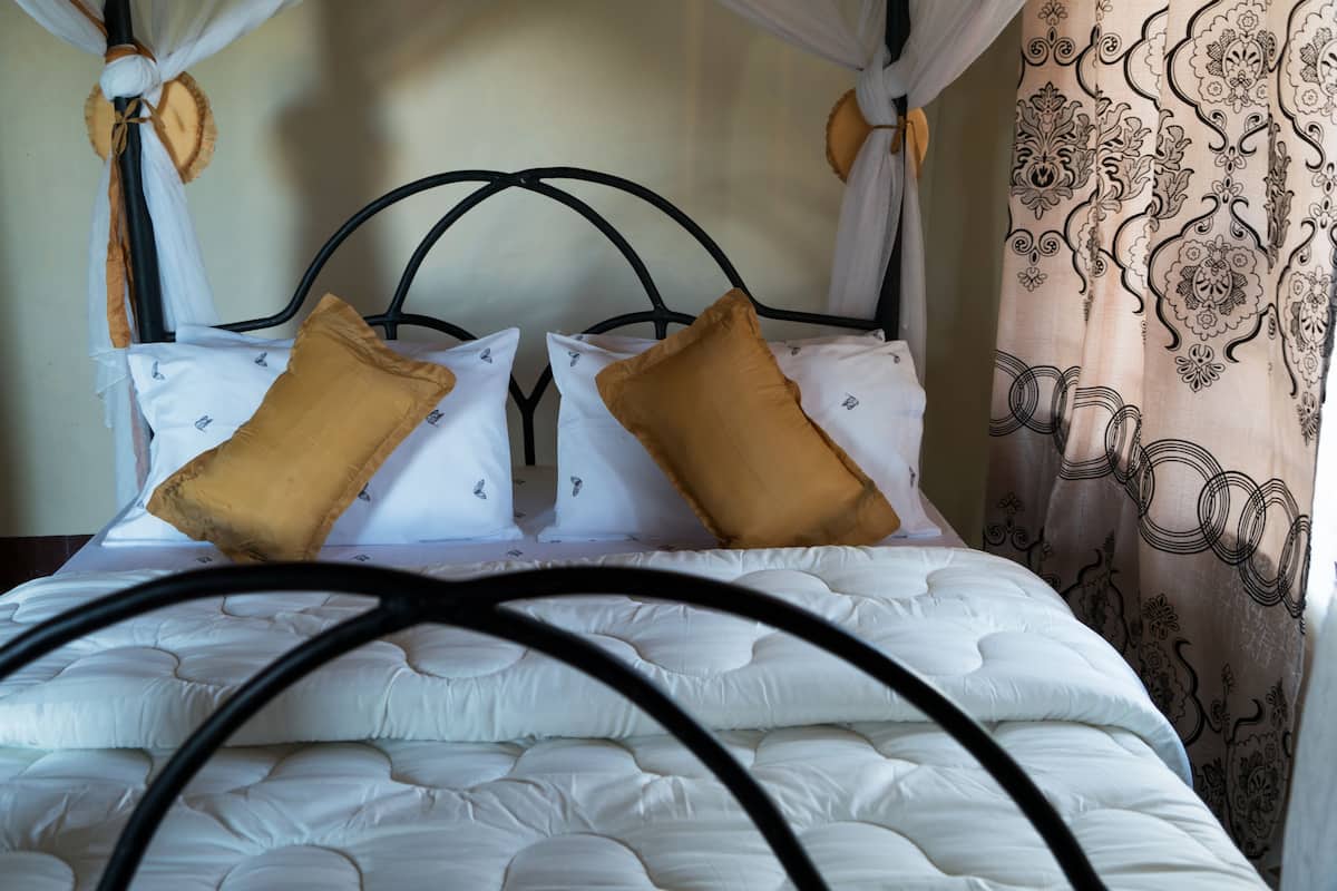 Uma cama com colcha branca e duas almofadas marrons, estrutura de metal preto e dossel com tecido branco e laranja.