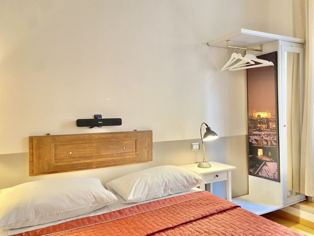 Quarto com cama, travesseiros, mesa de cabeceira com abajur, cabeceira de madeira e vista para a paisagem urbana pela janela.