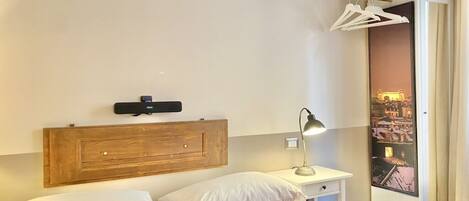 Quarto com cama, travesseiros, mesa de cabeceira com abajur, cabeceira de madeira e vista para a paisagem urbana pela janela.
