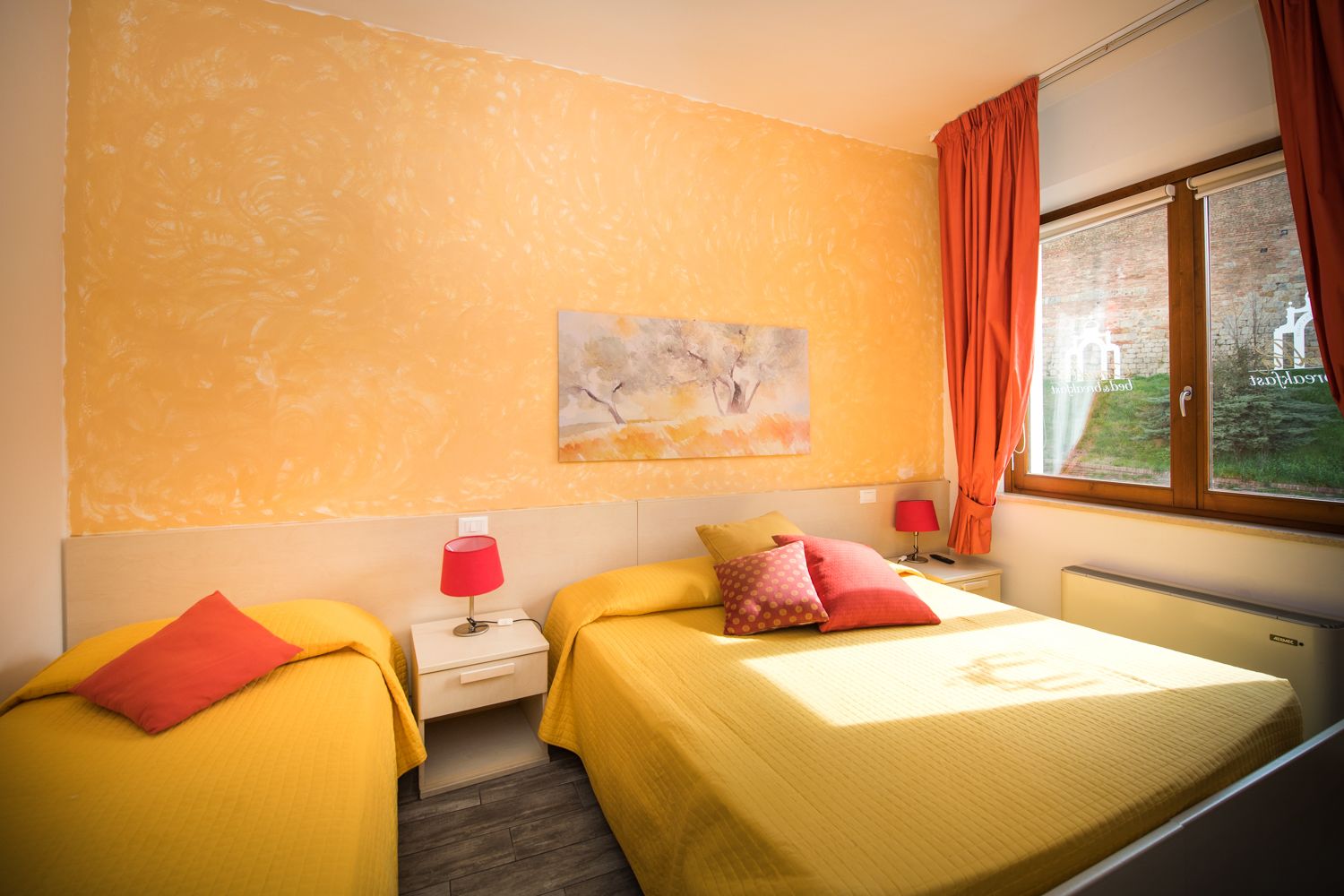Quarto de hotel com duas camas, cada uma com roupa de cama amarela e almofadas vermelhas, uma pequena mesa de cabeceira com abajur, um quadro na parede e uma janela com cortinas.