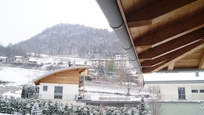 Um edifício moderno com fachada de madeira e telhado inclinado, situado em uma encosta nevada, com edifícios residenciais ao fundo.
