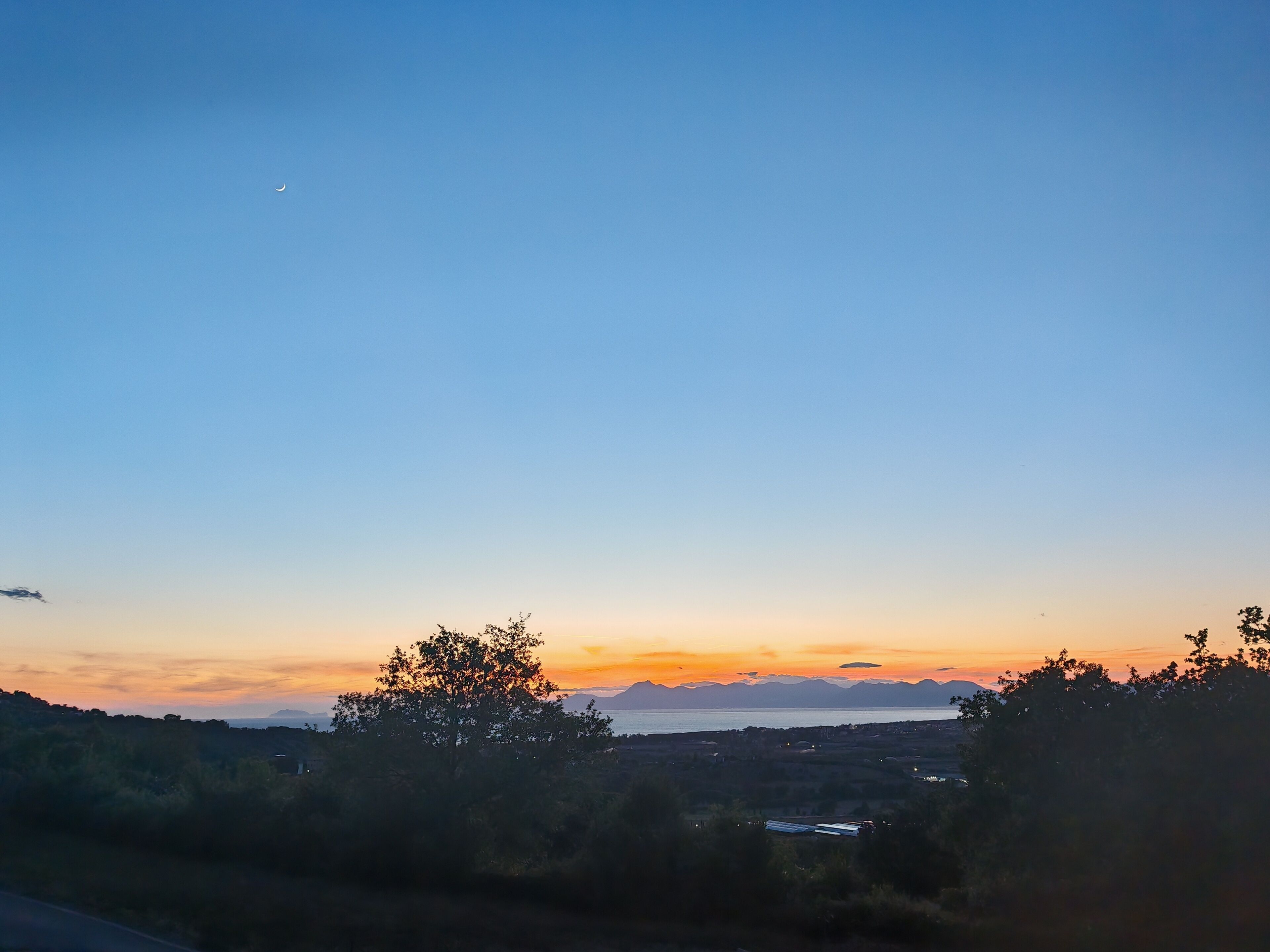 Vista do pôr do sol com montanhas ao longe e uma árvore em primeiro plano.
