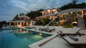 Piscina com espreguiçadeiras e área coberta, cercada por uma casa grande com vários andares e varandas.