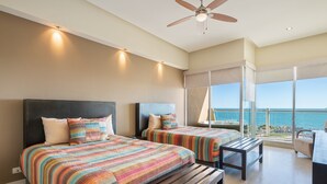 Quarto de hotel com duas camas, ventoinha no teto e uma grande janela com vista para o oceano.