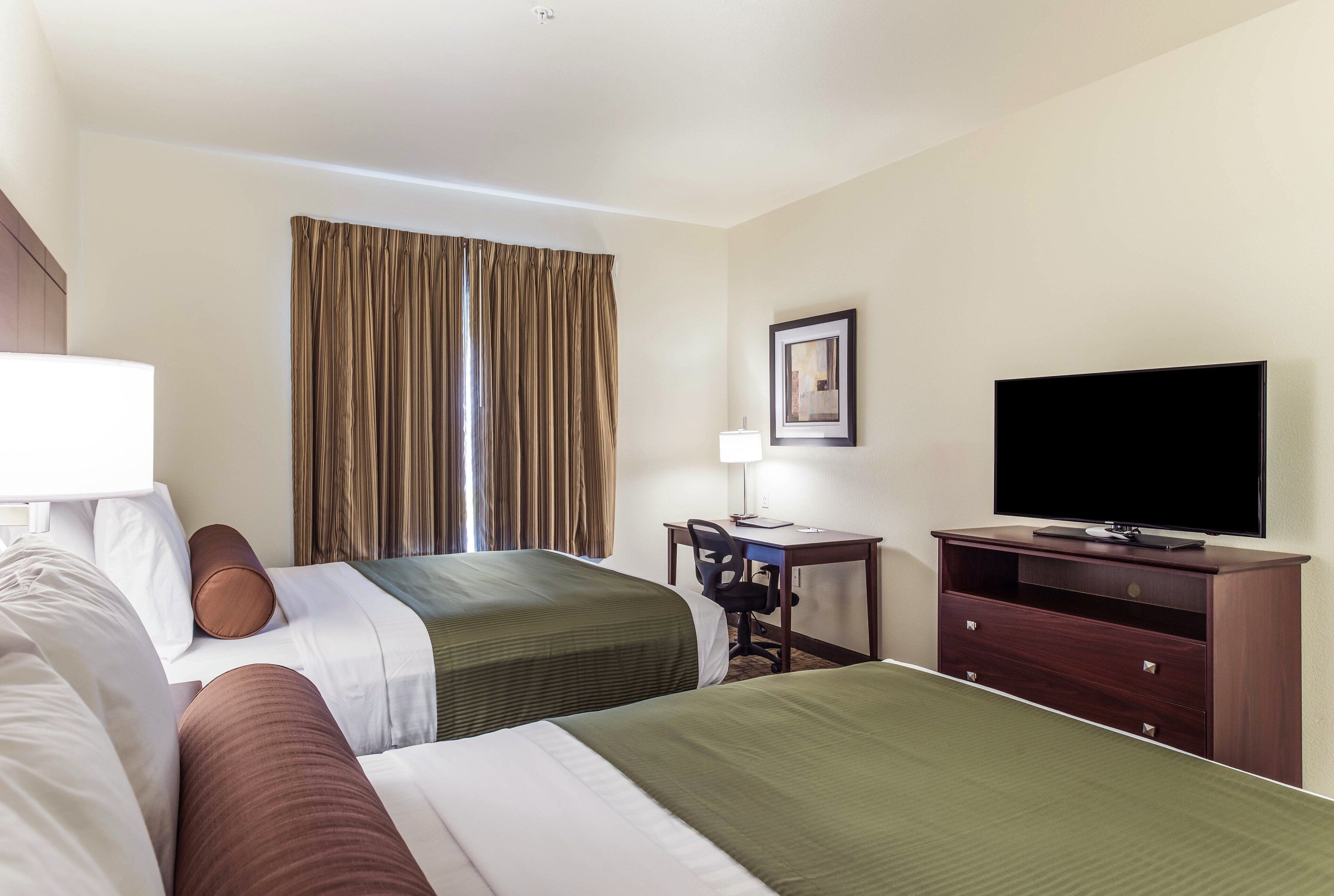 Quarto de hotel com duas camas, uma escrivaninha com cadeira, televisão e janela com cortinas.