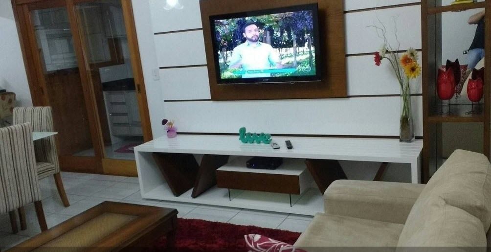 Uma sala de estar com uma televisão de tela plana presa a uma parede branca, um móvel branco para entretenimento, um sofá bege e um tapete vermelho.
