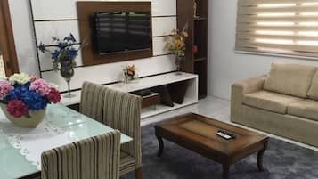 Uma sala de estar com sofá, mesa de centro e televisão na parede. Há uma mesa de jantar com cadeiras e um vaso de flores.