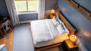 Uma cama bem arrumada com lençóis brancos, cabeceira de madeira e duas luminárias laterais. Uma janela com cortinas é visível, e uma pequena mesa com uma cadeira está no canto.