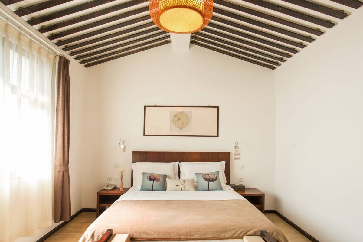 Quarto com teto de madeira, cama grande com várias almofadas, mesas de cabeceira e luminária pendente.