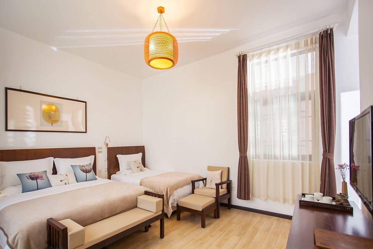 Quarto de hotel com duas camas, um banco de madeira, uma televisão e uma janela grande com cortinas.