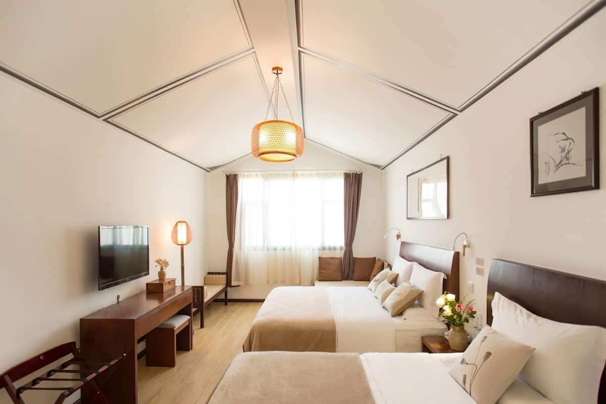 Quarto de hotel com duas camas, uma escrivaninha, televisão e uma janela grande com cortinas.