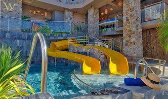 Uma área de piscina com um escorregador amarelo, paredes de pedra e um guarda-corpo de vidro.