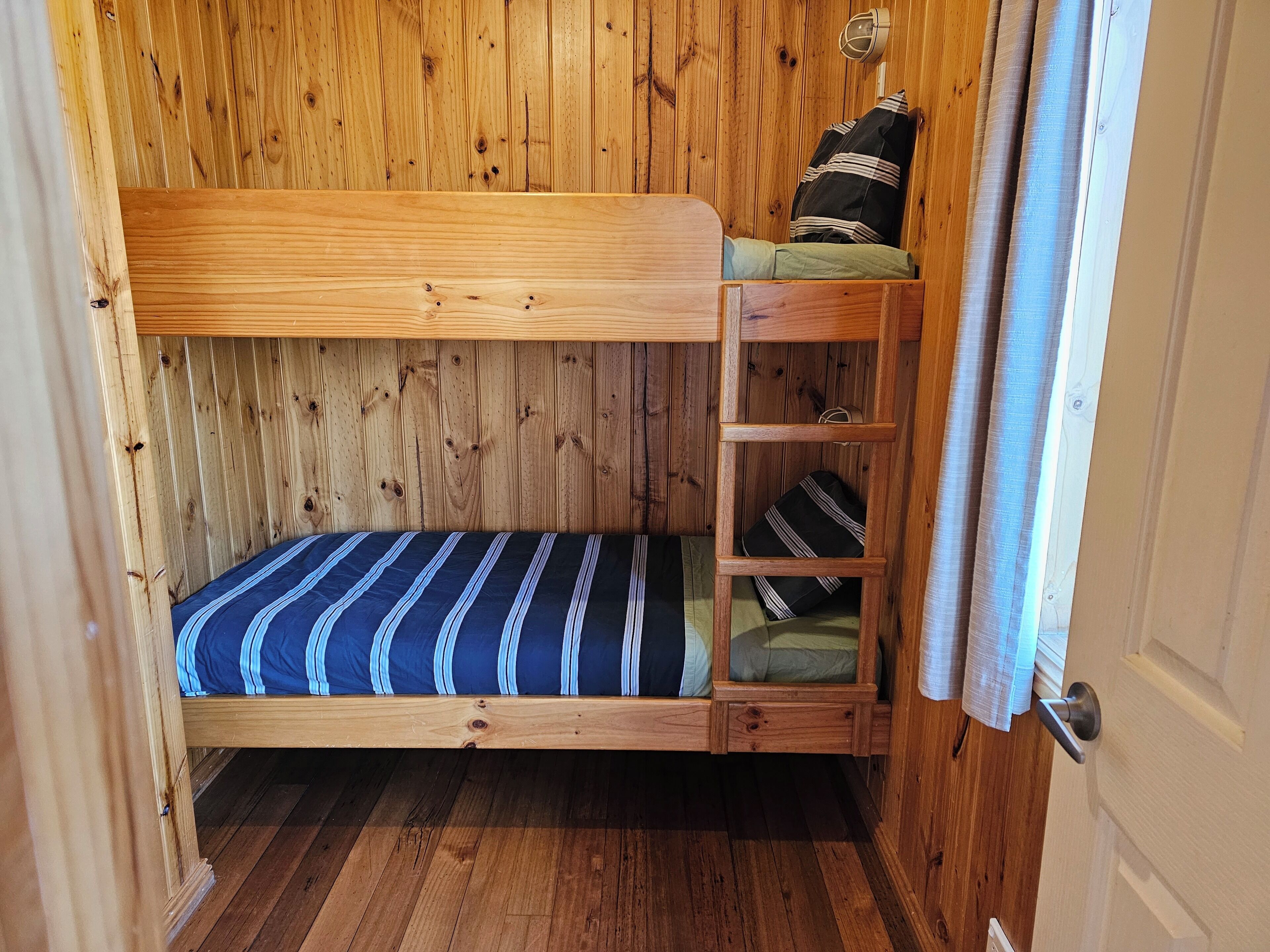 Uma cama beliche de madeira com escada, um pequeno armário e uma porta com cortina.