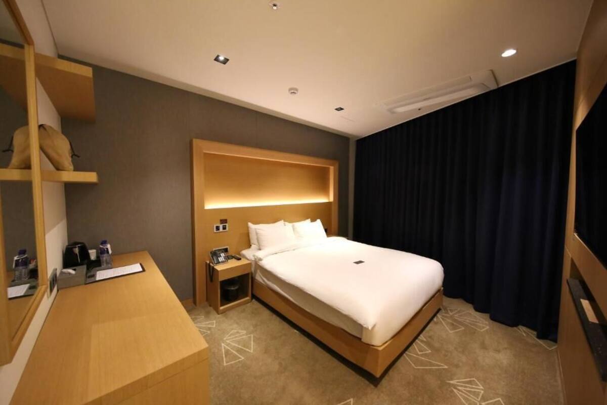 Quarto de hotel com uma cama grande, escrivaninha de madeira, mesa de cabeceira com abajur e cabeceira com iluminação embutida.