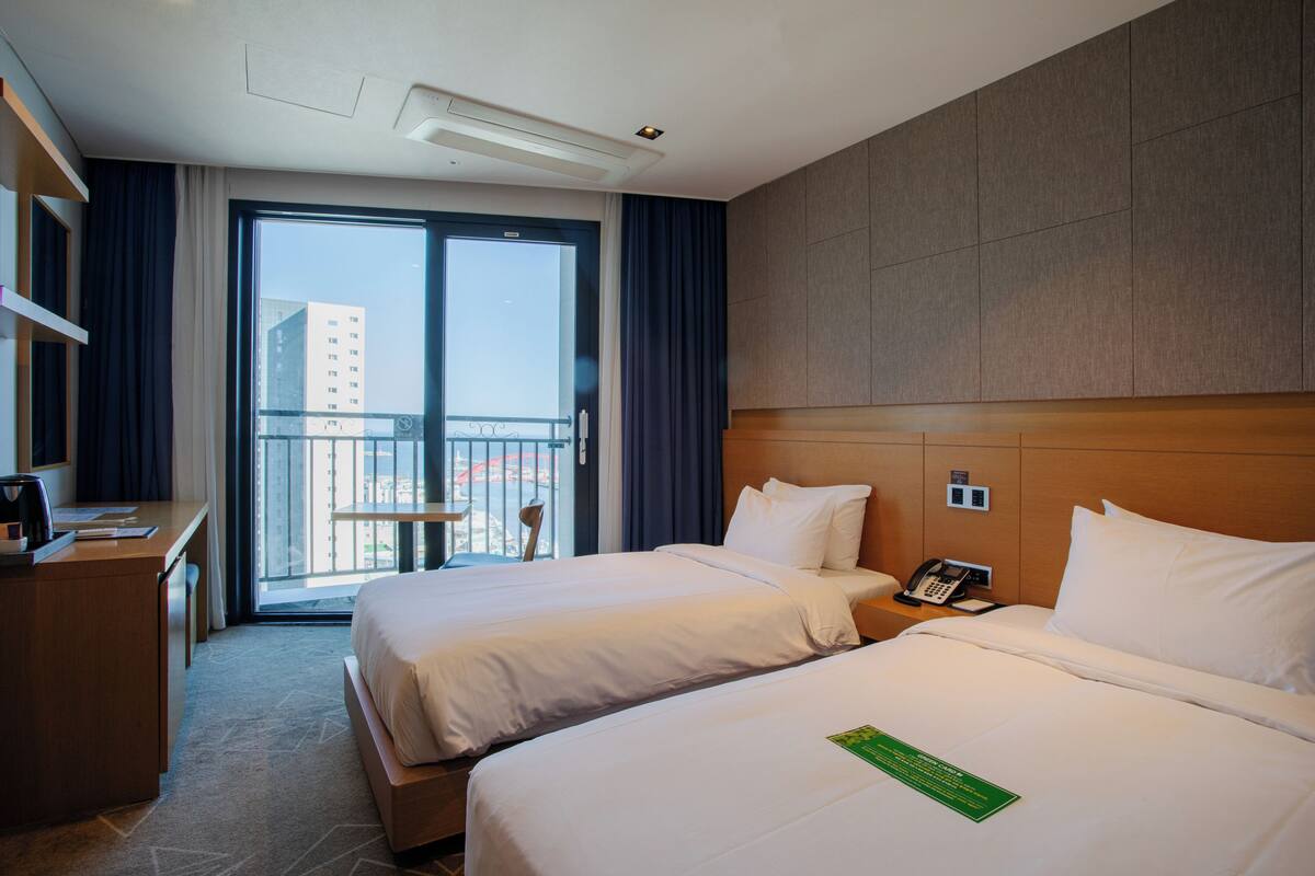 Quarto de hotel com duas camas, uma escrivaninha, uma cadeira, um telefone e janela com vista para edifícios.