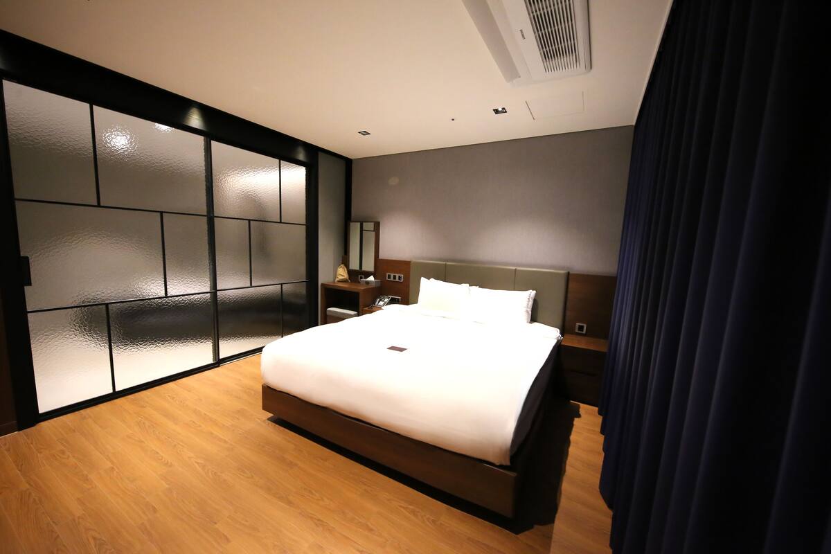 Quarto de hotel moderno com uma cama grande, piso de madeira, armário embutido e mesa de cabeceira.