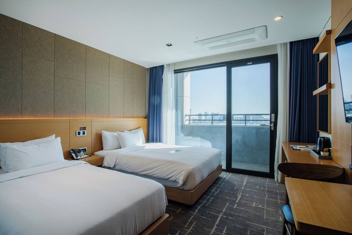Quarto de hotel com duas camas, uma mesa, uma cadeira, um telefone e vista da cidade através de grandes janelas.