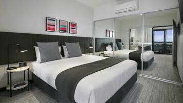 Quarto de hotel moderno com uma cama grande, uma mesa de cabeceira com abajur, uma mesinha lateral com revista e um espelho grande que reflete o quarto.