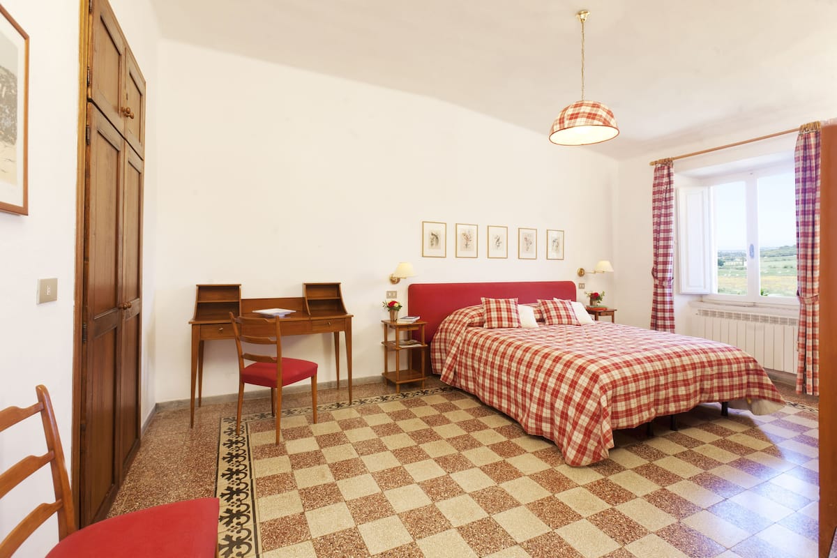 Quarto com piso xadrez, cama com colcha xadrez vermelha e branca, escrivaninha com cadeira, guarda-roupa de madeira e janela com cortinas.
