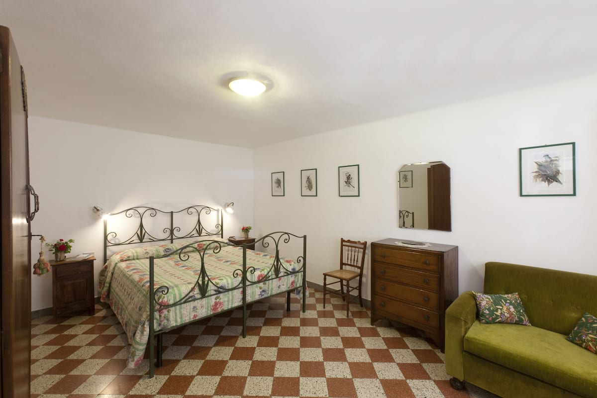 Um quarto com piso xadrez, uma cama com colcha floral, cômoda de madeira, sofá verde e um espelho na parede.