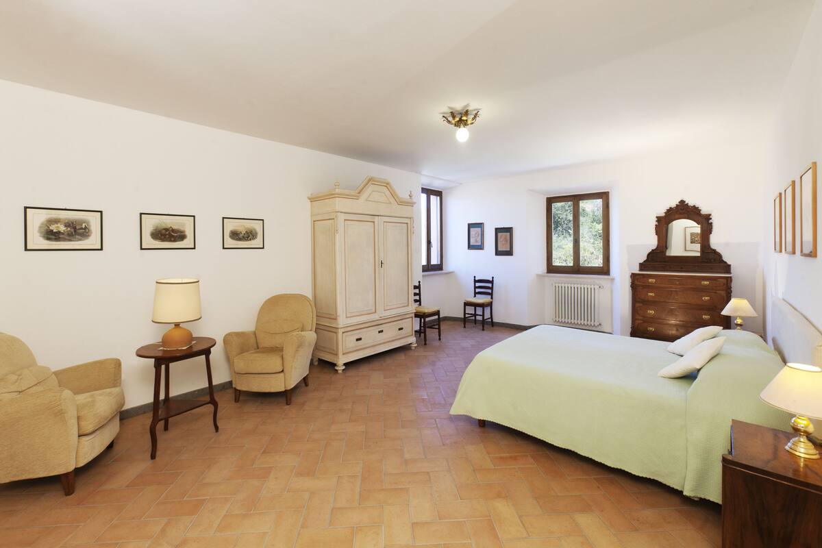 Um quarto com cama, cómoda, cadeira, candeeiro e armário.