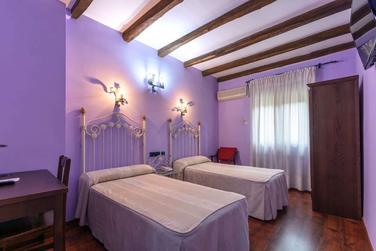Quarto com duas camas, uma mesa de madeira, uma cadeira e uma televisão na parede.