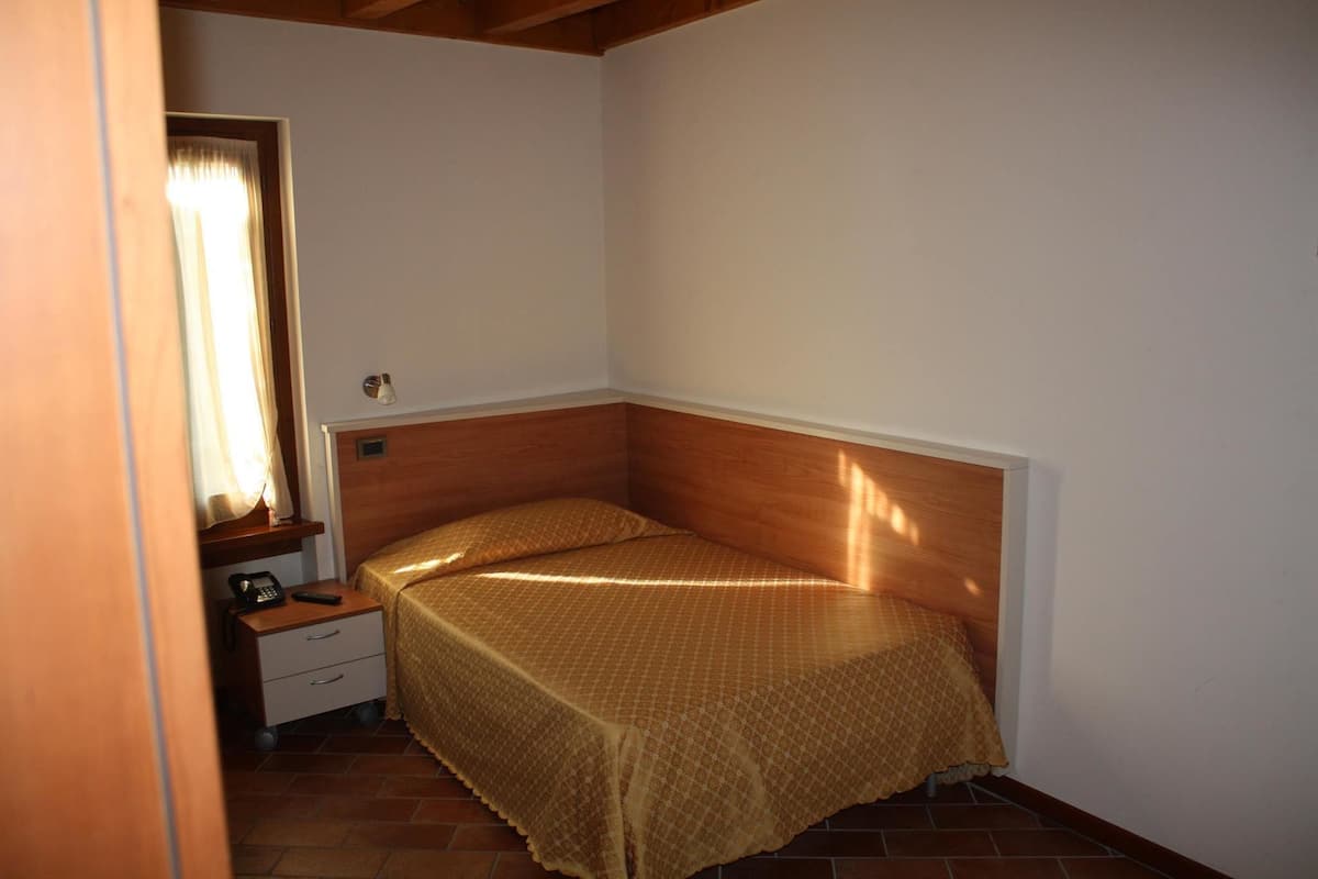 Quarto pequeno com cama de solteiro, cabeceira de madeira, mesa de cabeceira com abajur e janela com cortinas.