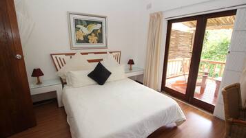 Quarto com cama, mesa de cabeceira, luminária, pintura e varanda com cadeira.