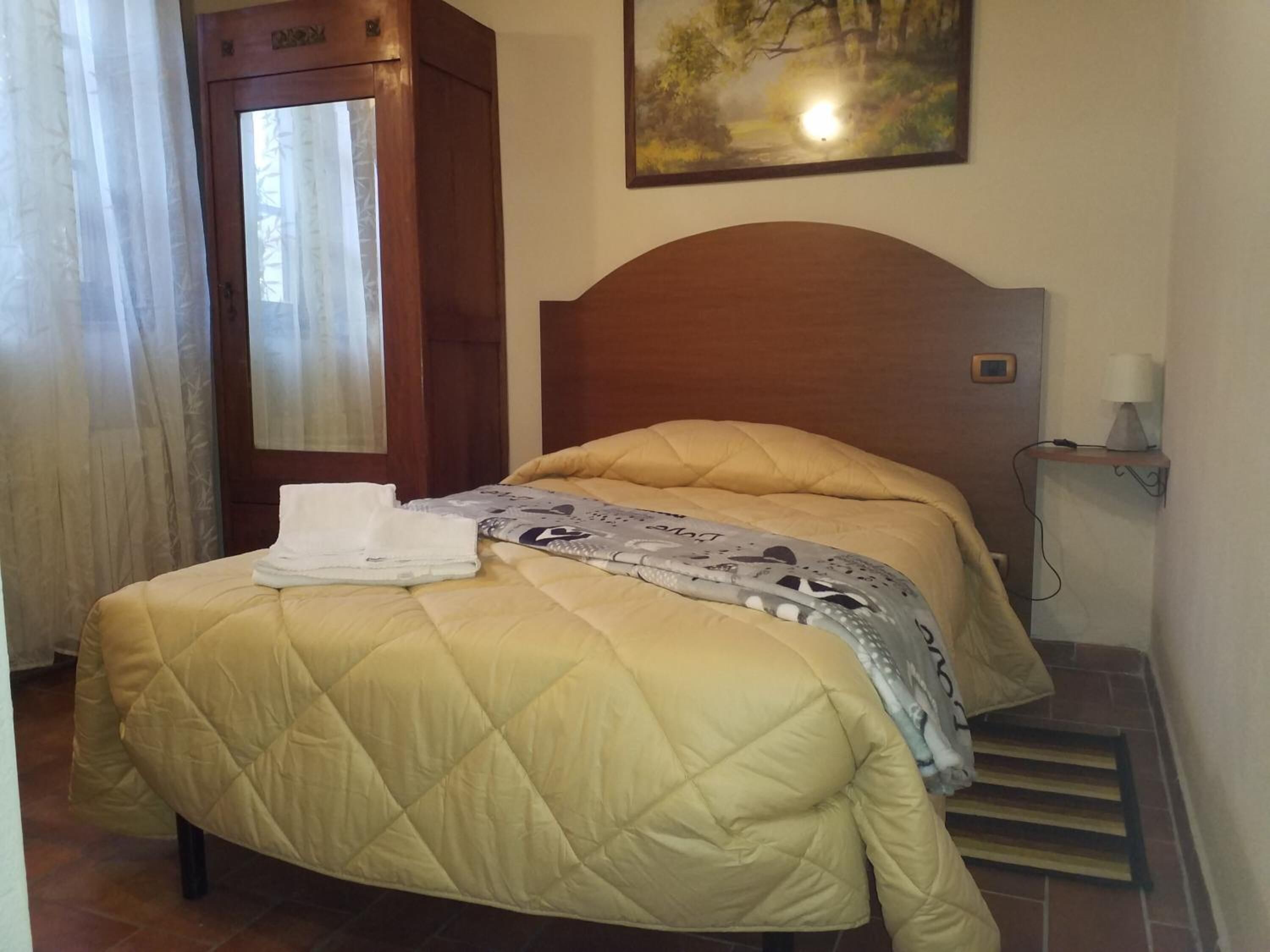 Cama bem arrumada com colcha amarela acolchoada e manta estampada, cabeceira de madeira, pintura de paisagem na parede, janela com cortinas finas e uma pequena mesa de cabeceira com abajur.