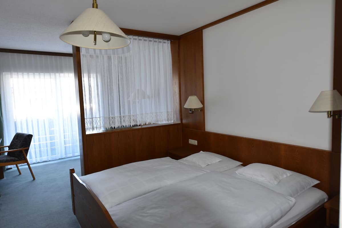 Quarto de hotel com cama de madeira, duas luminárias de cabeceira, uma cadeira e uma janela com cortinas leves.