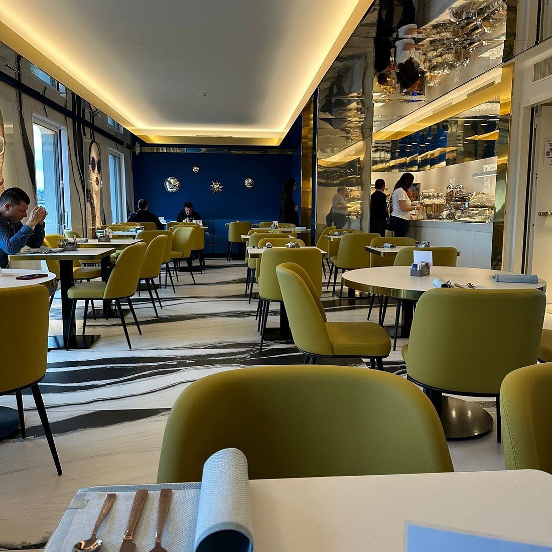 Uma área de jantar moderna com cadeiras e mesas amarelas, uma parede de destaque azul e vista para a cidade através de amplas janelas.