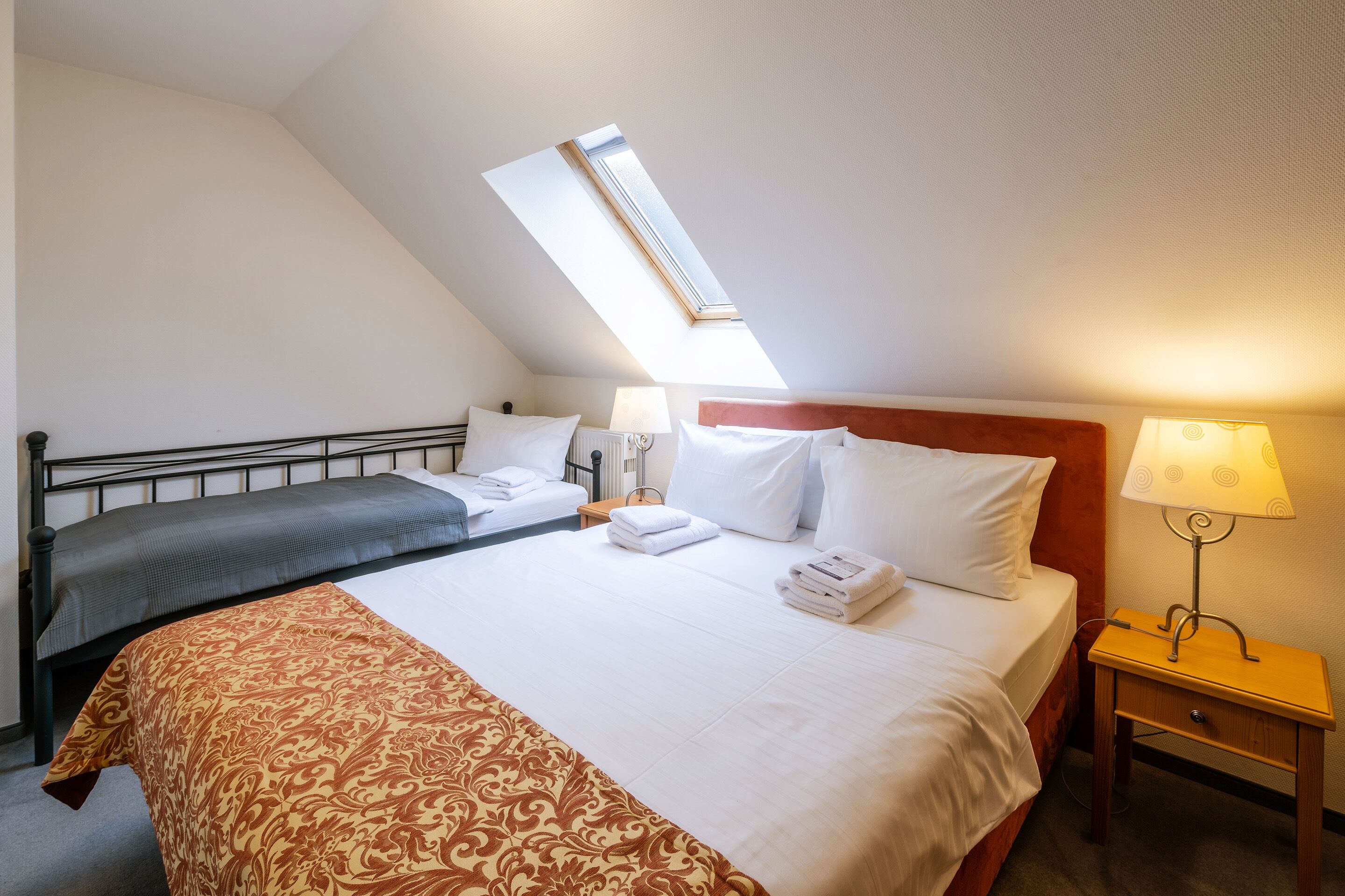 Quarto com duas camas, um claraboia, mesa de cabeceira com abajur e colcha estampada.