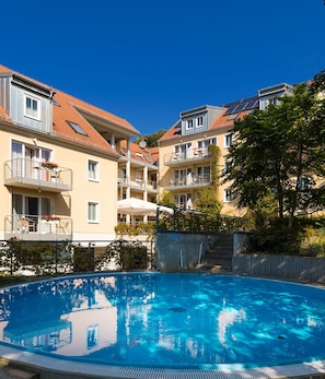 Piscina rodeada por edifícios altos com varandas, céu azul límpido e alguma vegetação.