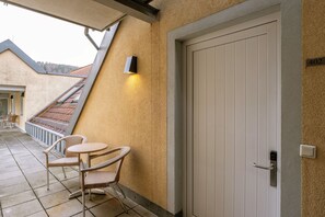 Uma área de estar coberta ao ar livre com cadeiras de vime e uma mesa, uma porta branca com placa de número e um edifício com telhado inclinado.
