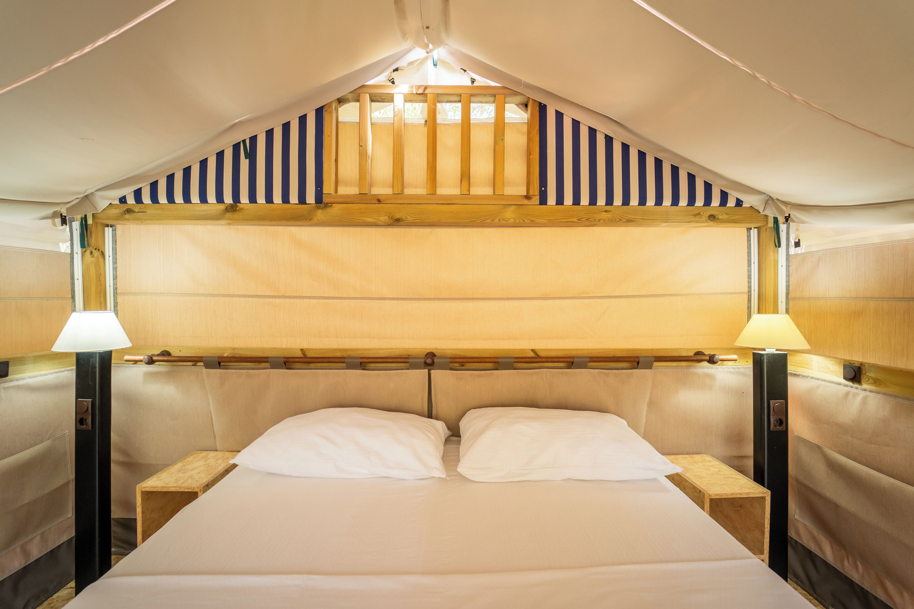Uma cama de solteiro com roupa de cama branca em um quarto tipo tenda, com cabeceira de madeira e duas luminárias de cabeceira.
