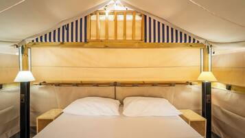 Uma cama de solteiro com roupa de cama branca em um quarto tipo tenda, com cabeceira de madeira e duas luminárias de cabeceira.