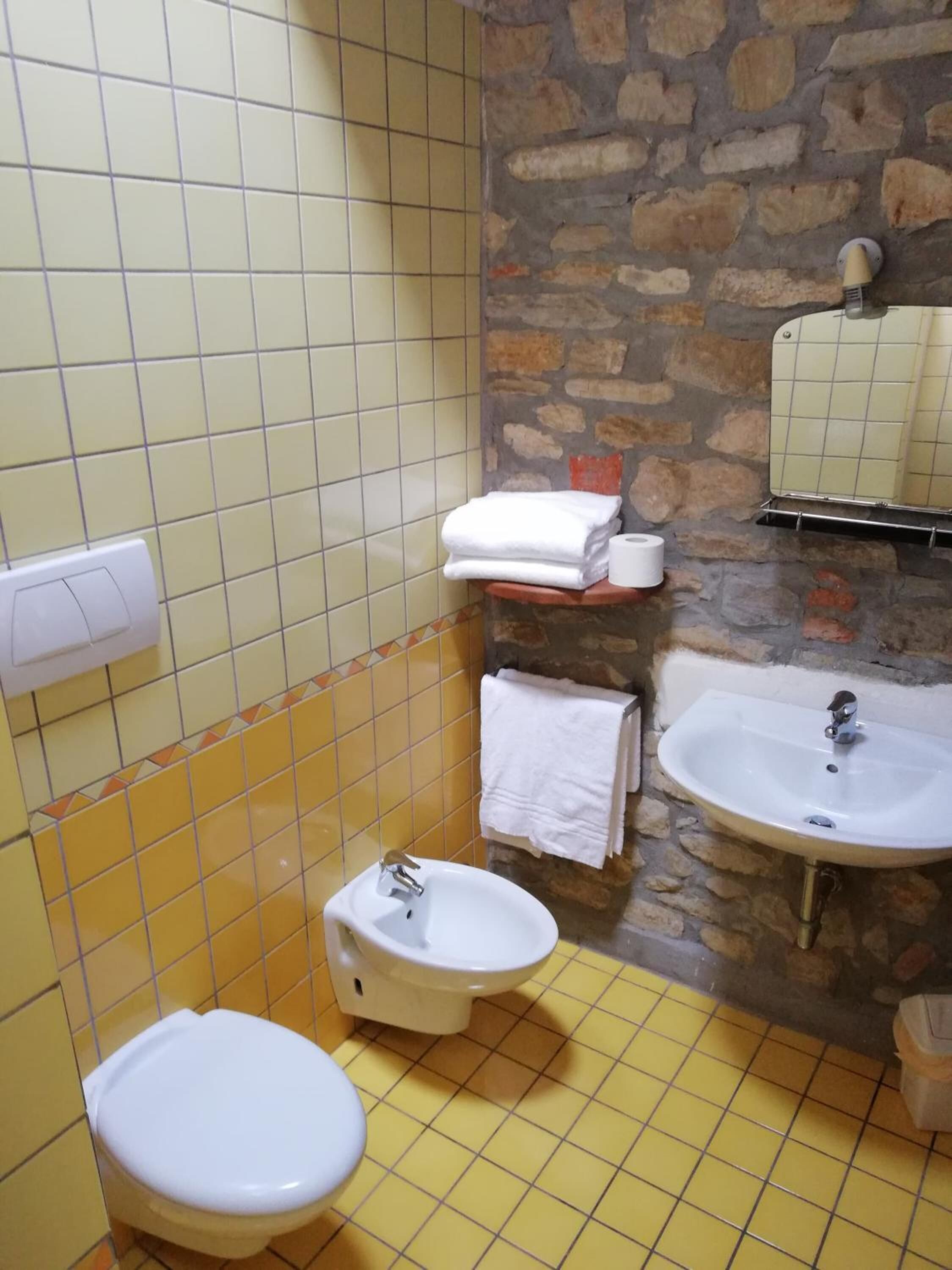 Casa de banho com paredes revestidas a azulejos amarelos, uma parede de destaque em pedra, uma sanita branca, uma sanita suspensa e um bidé.