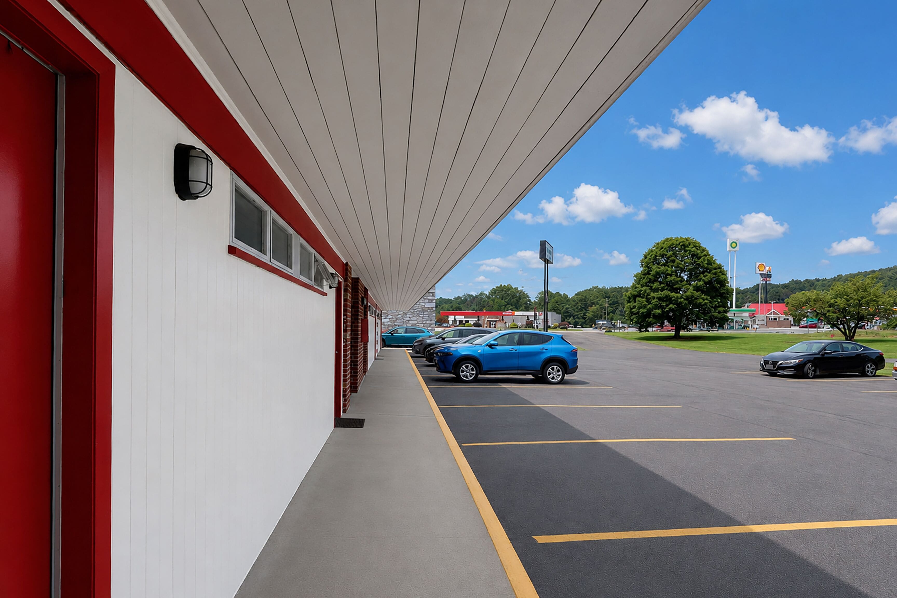 A covered walkway leading to a building with a red and white facade, a parking lot with several cars, and a clear blue sky with clouds.