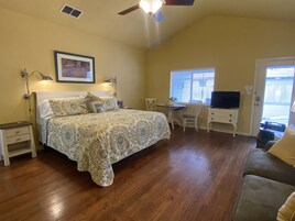 Um quarto com cama, mesas de cabeceira, escrivaninha, cadeira, televisão e janela com persianas.
