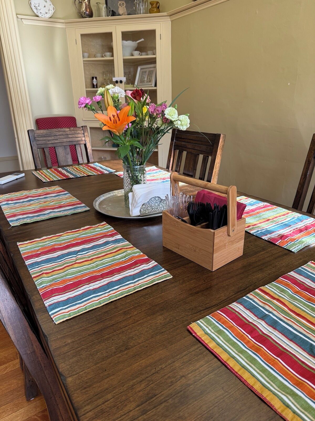 Uma mesa de jantar com jogos americanos listrados, um vaso com flores e um suporte de utensílios de madeira.