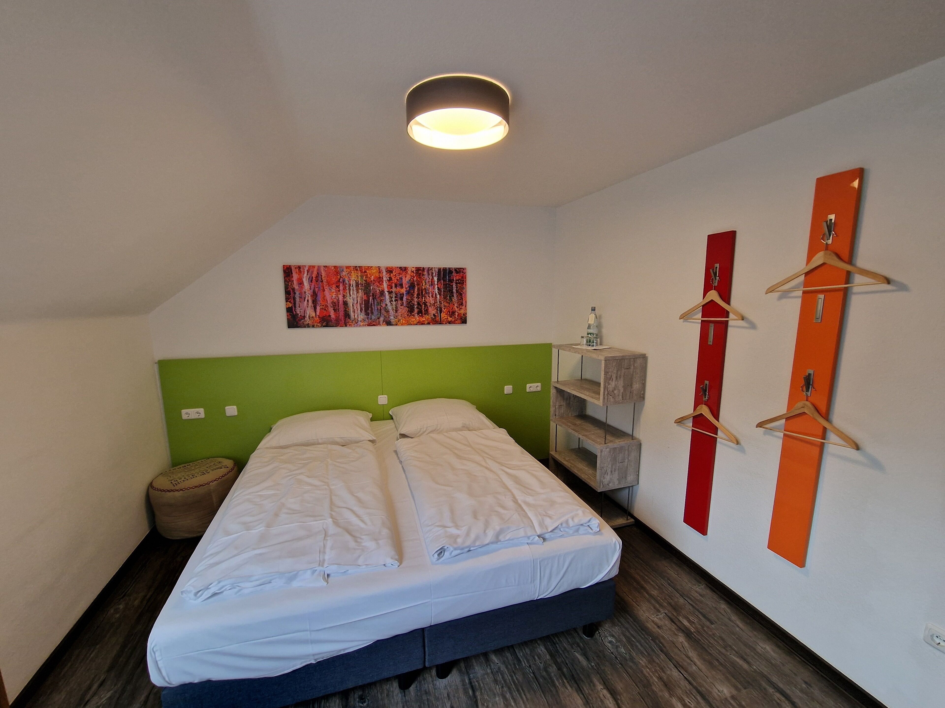 Cama bem arrumada em quarto com cabeceira verde, uma pequena prateleira com uma garrafa e um suporte na parede com cabides.
