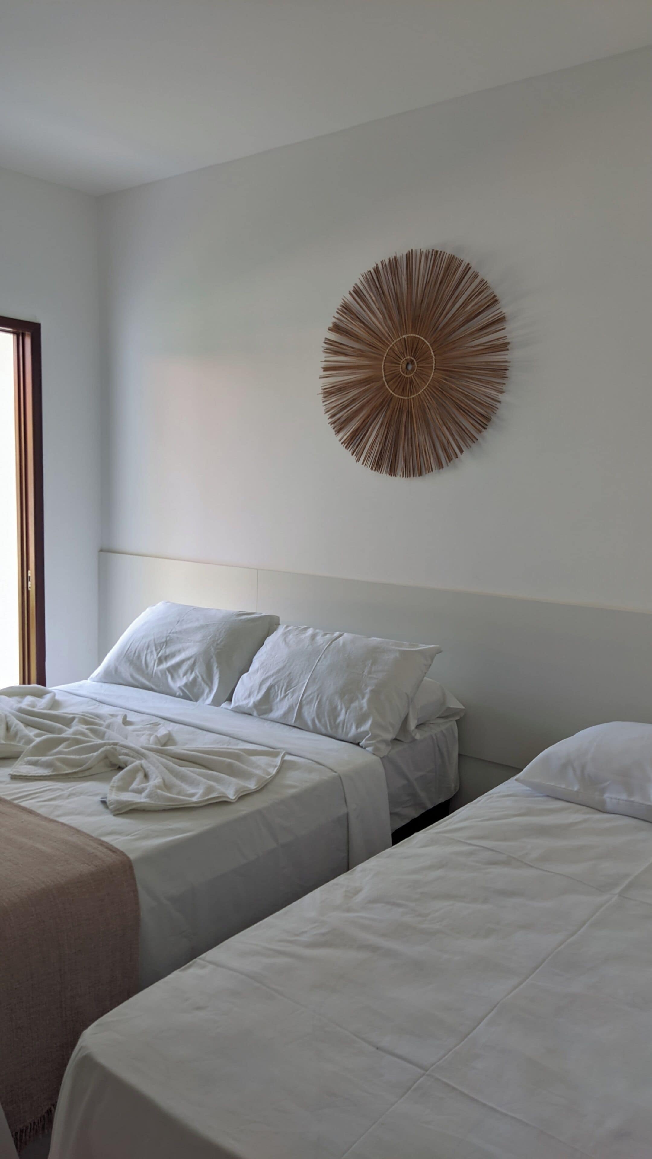 Quarto de hotel com duas camas, uma grande decoração circular na parede e uma janela à esquerda.