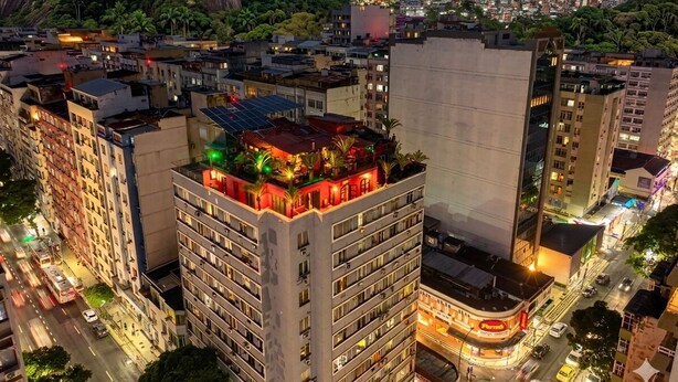 Um edifício alto com vários andares, um jardim no terraço e vista para a paisagem urbana com uma montanha imponente ao fundo.