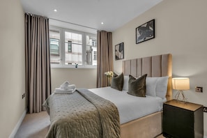 Quarto de hotel com cama, mesa de cabeceira, luminária e janela com cortinas.