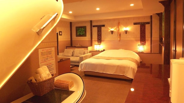 Quarto de hotel com cama, escrivaninha com luminária, cadeira, mesinha de centro com cesta e prateleira fixada na parede.