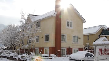 Um edifício de dois andares com chaminé de tijolos, várias janelas e um carro coberto de neve estacionado na frente.