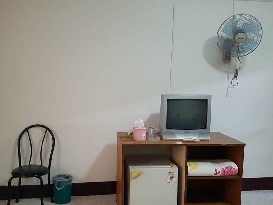 Quarto com escrivaninha de madeira, televisão, cadeira, lixeira e ventoinha fixada na parede.
