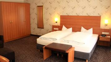 Quarto de hotel com cama de casal, guarda-roupa de madeira, um banco e uma mesa de cabeceira com luminária.